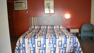 Quarto de hotel com colcha estampada, luminária de cabeceira, uma cadeira e ar condicionado na parede.