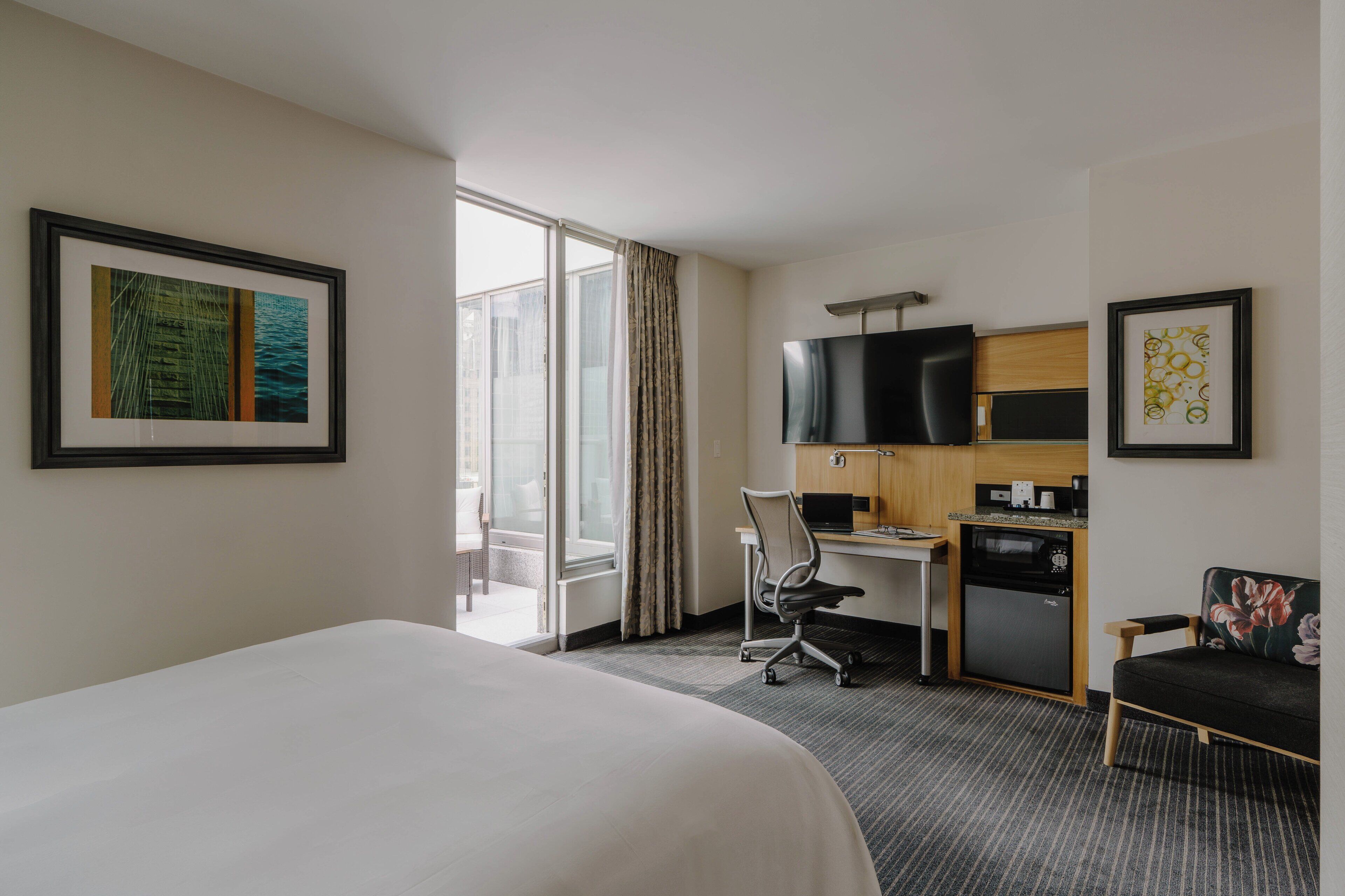 Quarto de hotel com cama, escrivaninha com computador, televisão, pequeno espaço de cozinha e sofá.