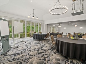 Sala de conferência com mesas, cadeiras, um quadro branco e uma janela grande com vista para o verde.