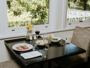 Mesa posta com pratos, copos e talheres, um vaso com uma flor, e vista para o verde pela janela.