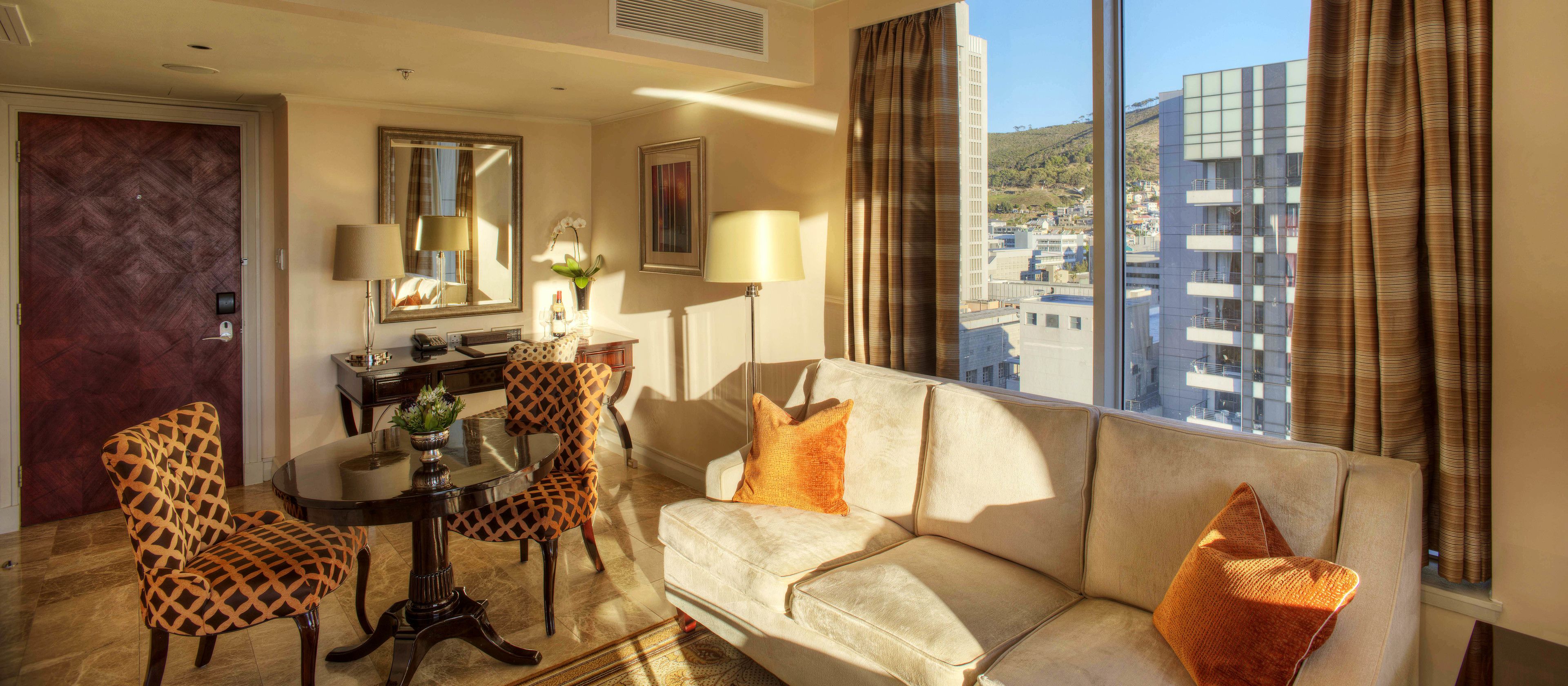 Quarto de hotel com sofá, poltronas, mesa de jantar e vista da cidade.