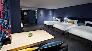 Quarto de hotel com duas camas, uma mesinha com uma planta e um mural colorido na parede esquerda.