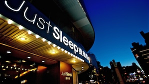 Placa de hotel com o nome 'Just Sleep' iluminado, telhado curvo e paisagem urbana ao fundo.