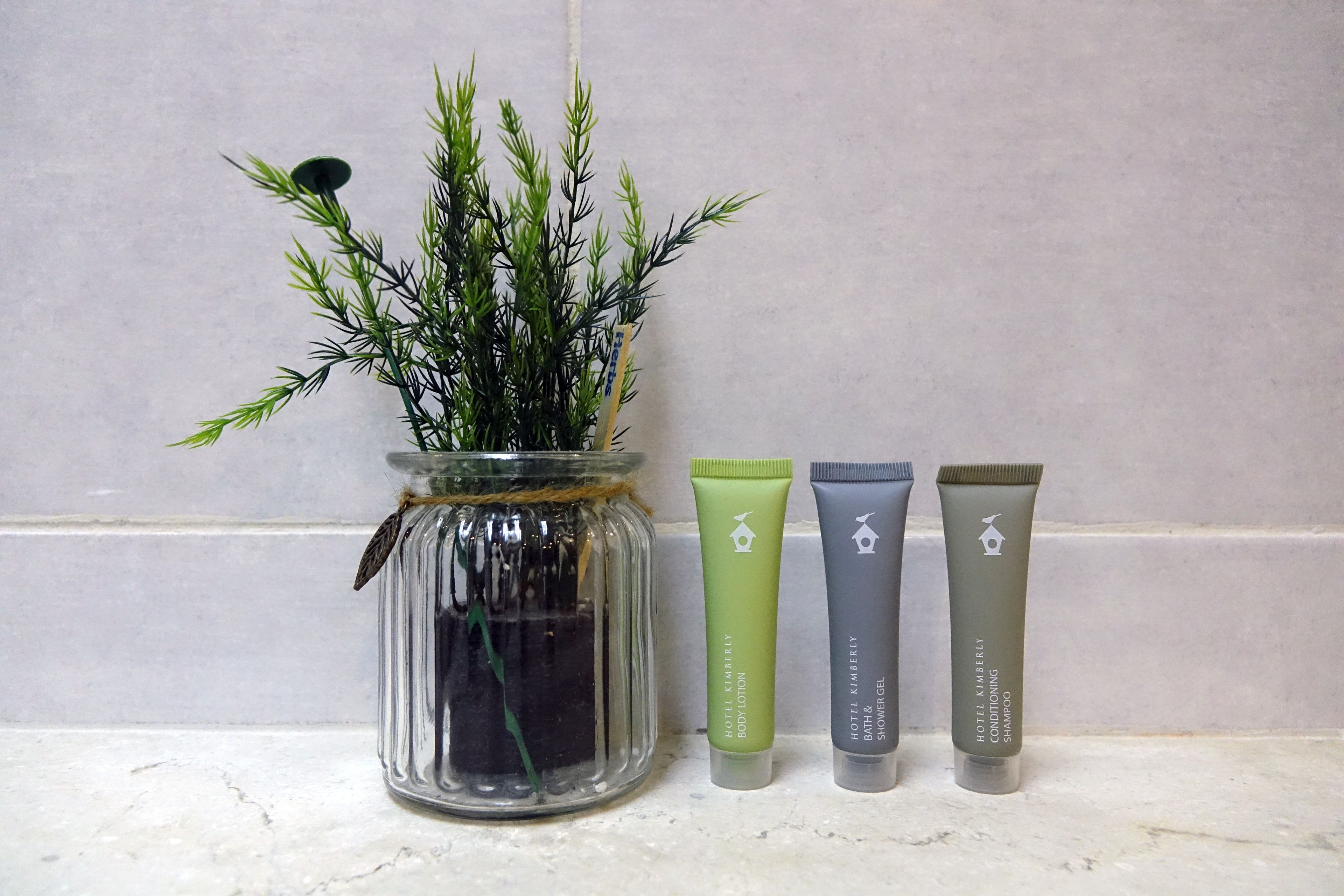 Um frasco de vidro com uma planta e três tubos de loção corporal, sérum para sobrancelhas e shampoo do Hotel Kimberly.