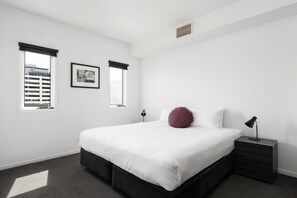 Quarto com cama, mesa de cabeceira, abajur, janela e quadro na parede.