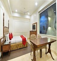 Quarto de hotel com cama, escrivaninha, cadeira, televisão e uma grande janela com vista para uma construção de pedra.