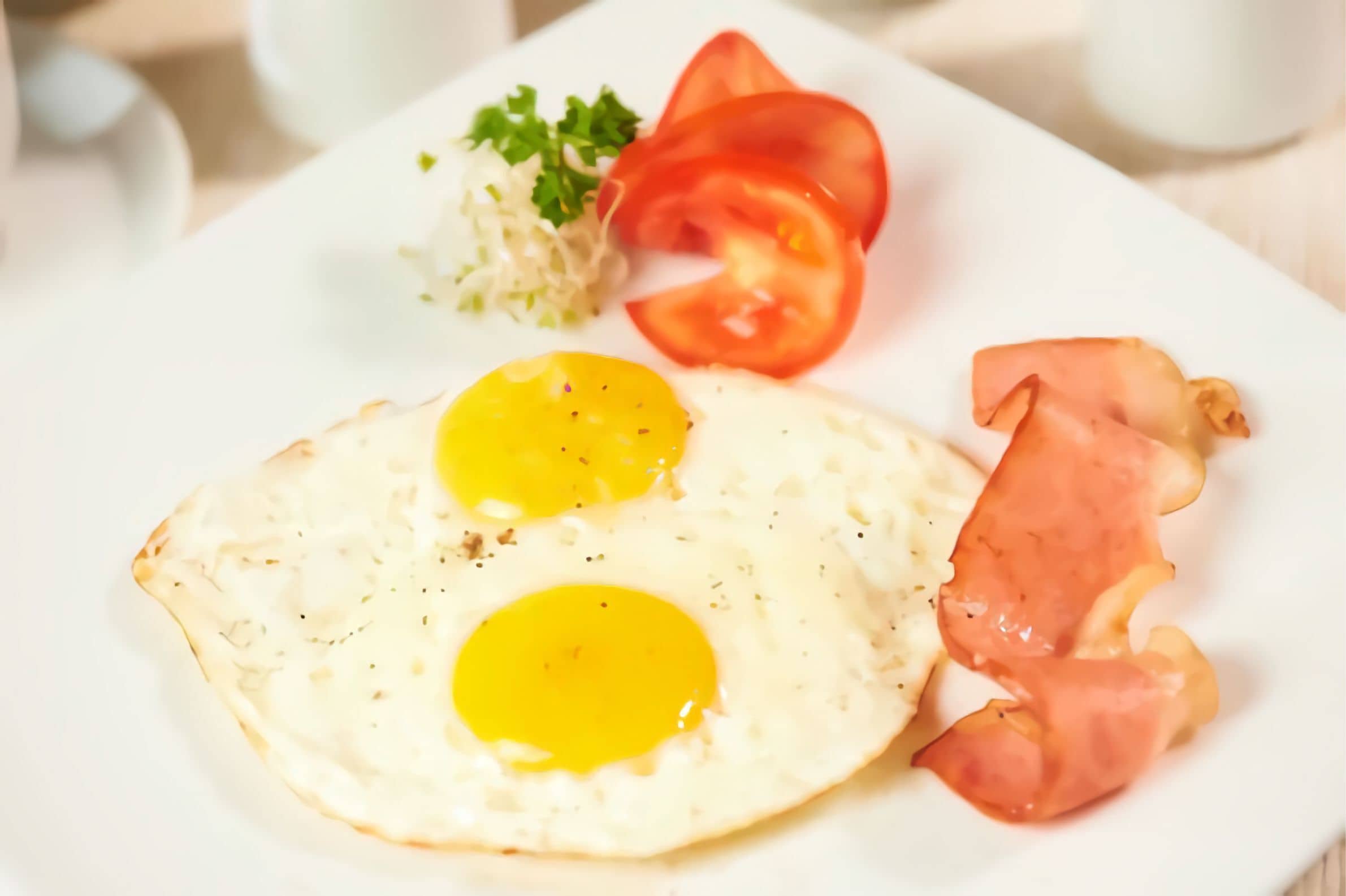 Um prato com dois ovos estrelados, uma fatia de tomate e um pedaço de presunto.