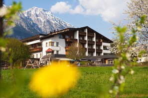 Um hotel com vários andares, varandas, um teleférico e montanhas ao fundo.