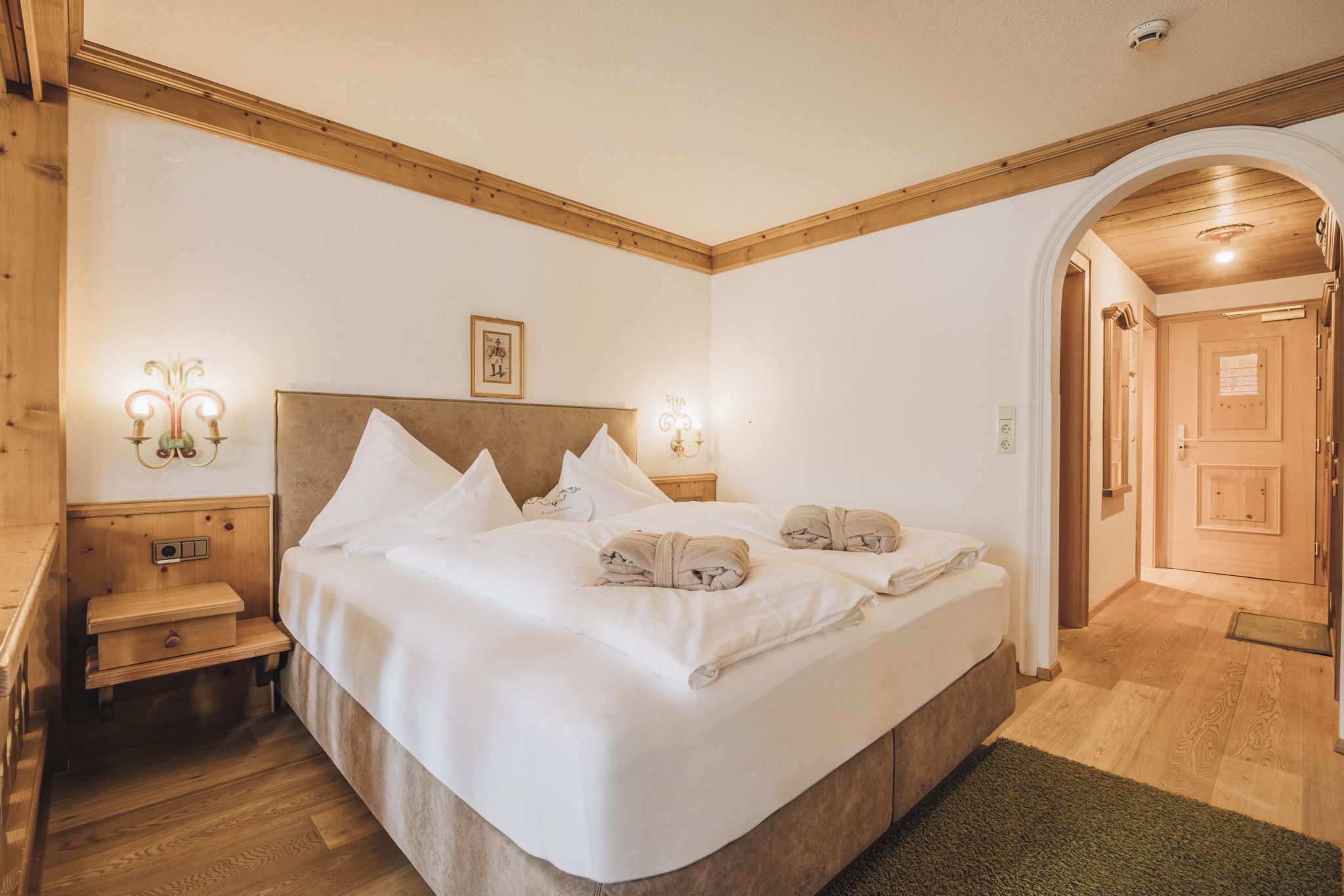 Quarto de hotel com uma cama grande, cabeceira de madeira, duas mesas de cabeceira, um criado-mudo e um candeeiro fixado na parede, além de uma porta que dá para outro cômodo.