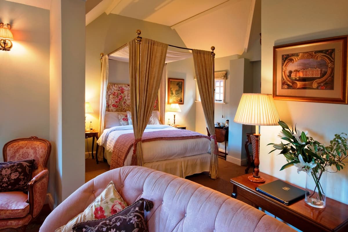 Um quarto com cama de dossel, um espaço de estar com poltrona e sofá, e uma mesinha de cabeceira com abajur e um vaso de flores.