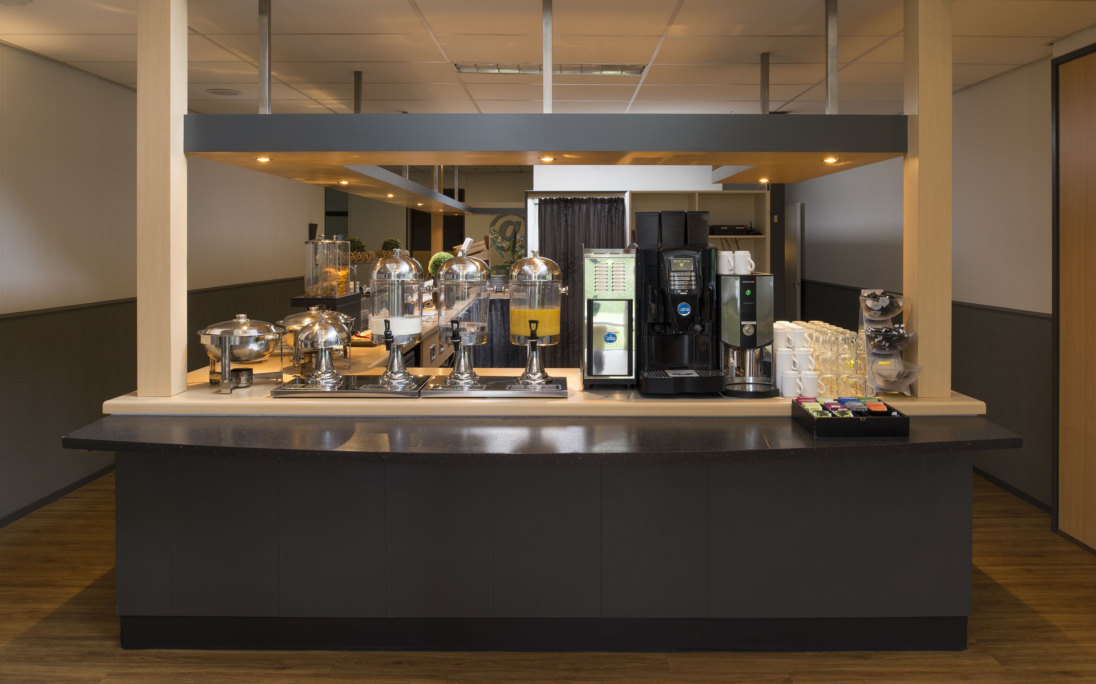 Uma área de recepção de hotel moderna, com um espaço para café à disposição, incluindo máquina de café, vaporizador de leite e diversos dispensadores de bebidas.