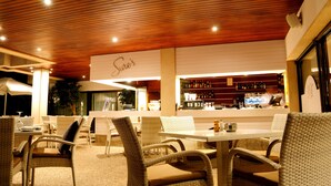 Uma área de estar externa com cadeiras e mesas de vime, um bar com prateleiras cheias de garrafas, e uma placa com a inscrição "Sure's".