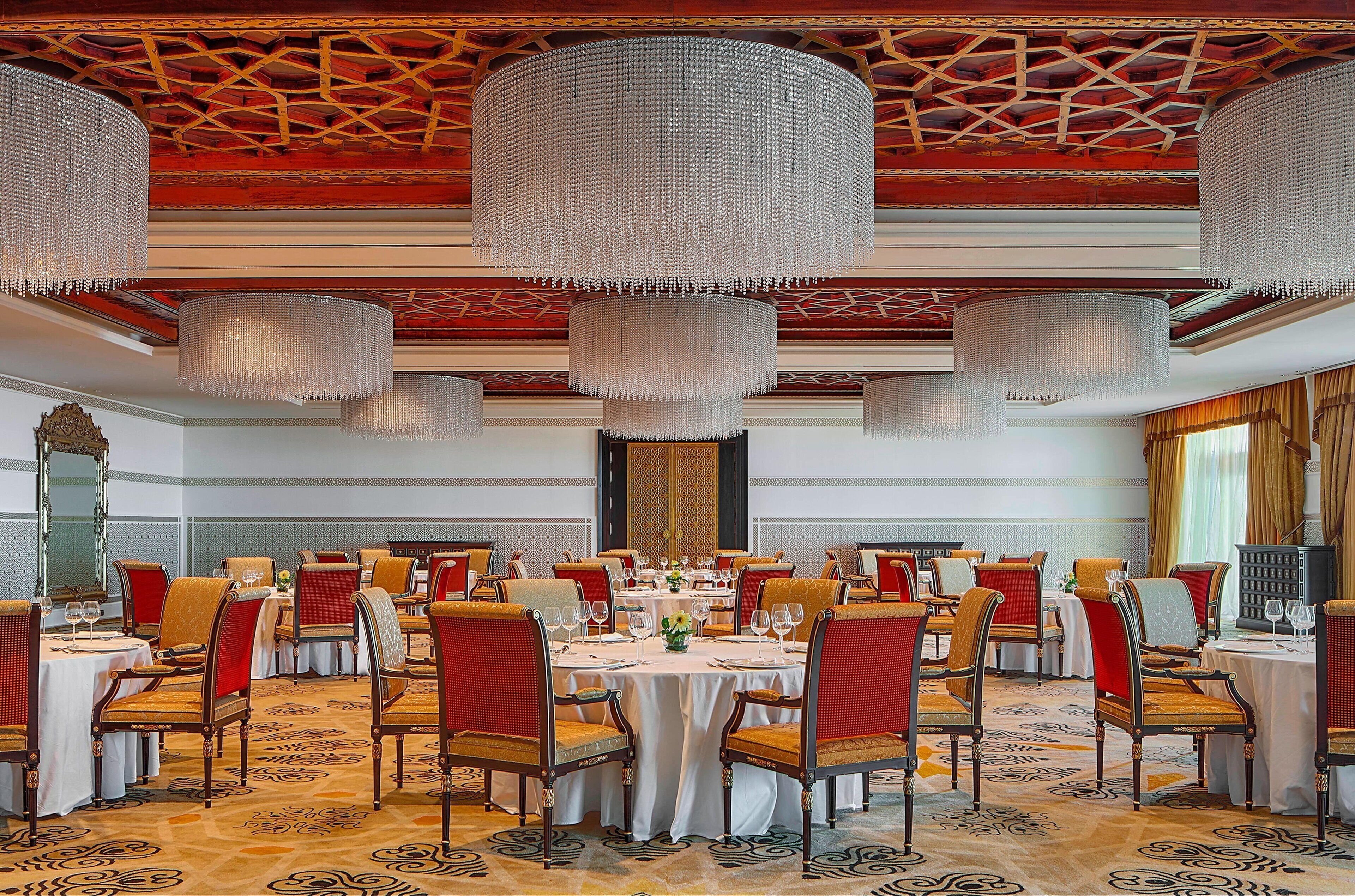 Uma sala de banquetes com várias mesas redondas, cadeiras estofadas em vermelho e grandes lustres de cristal.