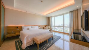 Quarto de hotel moderno com uma cama grande, um banquinho, uma televisão e vista para a cidade pela janela.