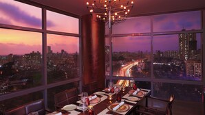 Uma sala de jantar com mesa posta para quatro pessoas, grandes janelas com vista para a cidade e um lustre moderno.