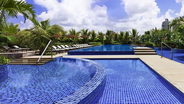 Uma piscina com azulejos azuis em mosaico, rodeada por palmeiras e cadeiras de descanso.