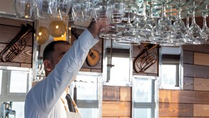 Um bartender organiza copos em um bar com parede de madeira e diversas garrafas ao fundo.