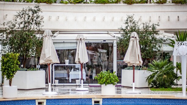 Piscina com espreguiçadeiras e guarda-sóis, cercada por vegetação e fachada de um edifício.