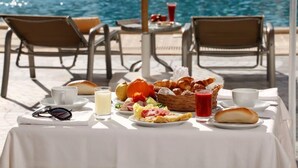 Mesa de café da manhã com croissants, frutas e suco à beira de uma piscina, com cadeiras e uma mesa.