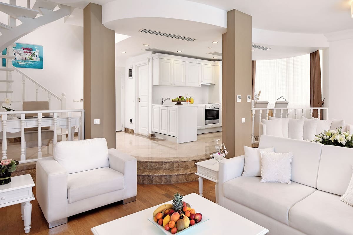 Uma sala de estar moderna com um sofá branco, uma mesa de centro com frutas e uma escada que dá para uma cozinha.