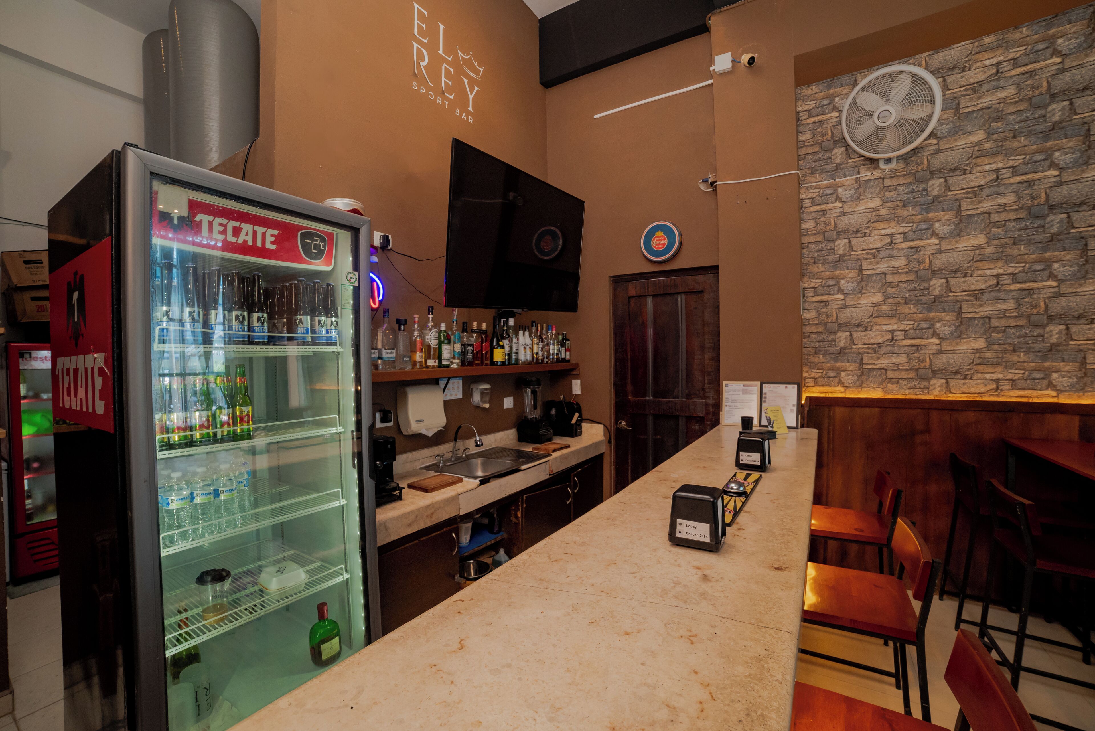 Uma área de bar com um refrigerador cheio de bebidas, uma TV de tela plana na parede e uma parede com textura de pedra.