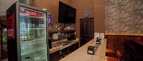 Uma área de bar com um refrigerador cheio de bebidas, uma TV de tela plana na parede e uma parede com textura de pedra.