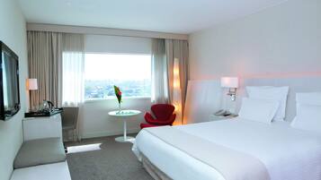 Quarto de hotel com uma cama grande, uma cadeira vermelha, uma mesinha e vista para a cidade pela janela.