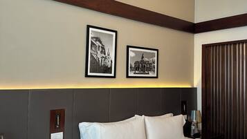 Quarto de hotel com uma cama grande, uma mesinha de cabeceira com telefone e abajur, e duas fotografias emolduradas na parede.