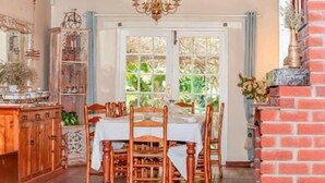 Uma área de refeições com mesa e cadeiras de madeira, um lustre e vista pela janela para o verde lá fora.