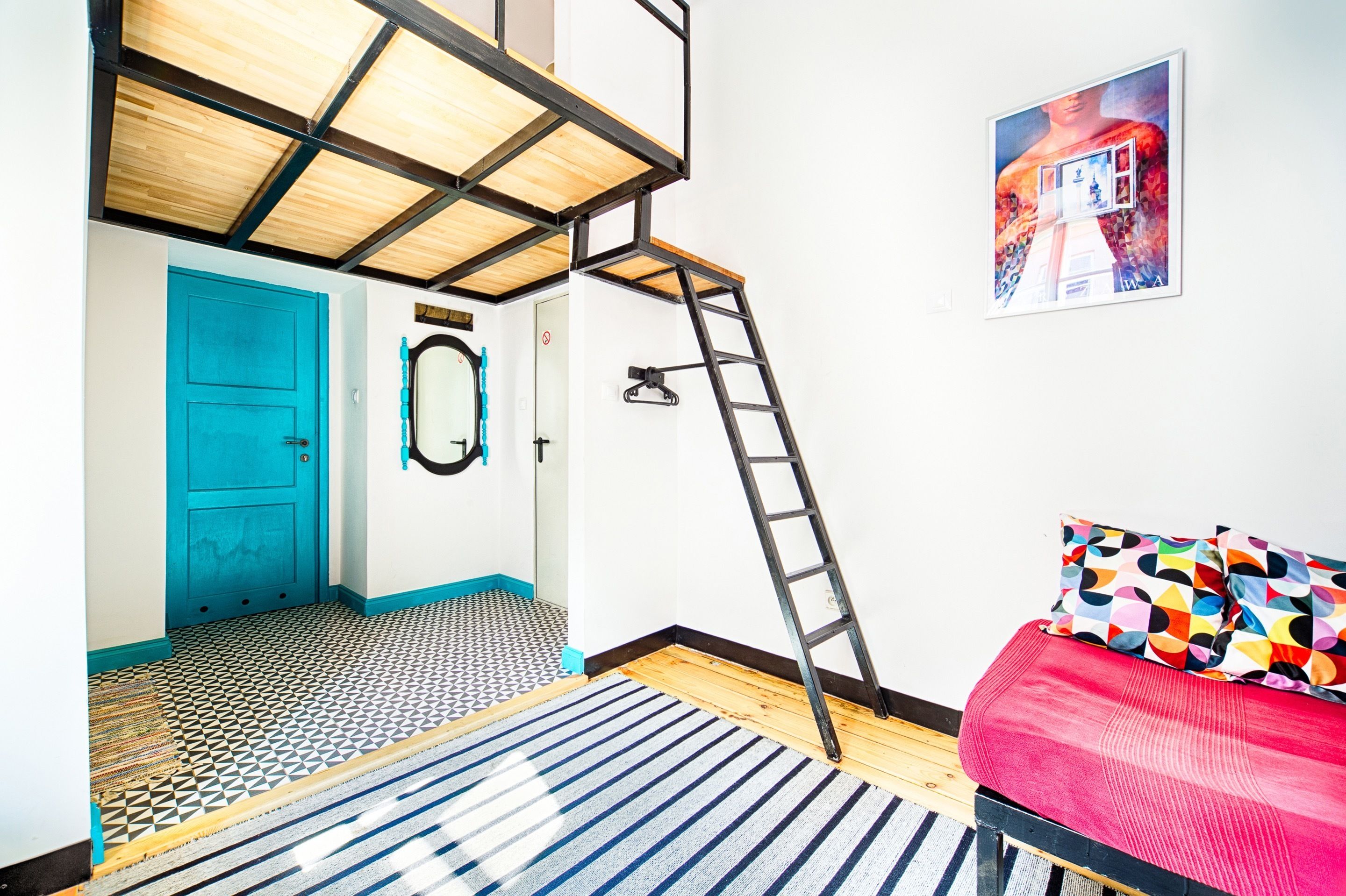 Um quarto com uma cama beliche, uma escada, uma porta azul, um espelho e uma cama com almofadas coloridas.
