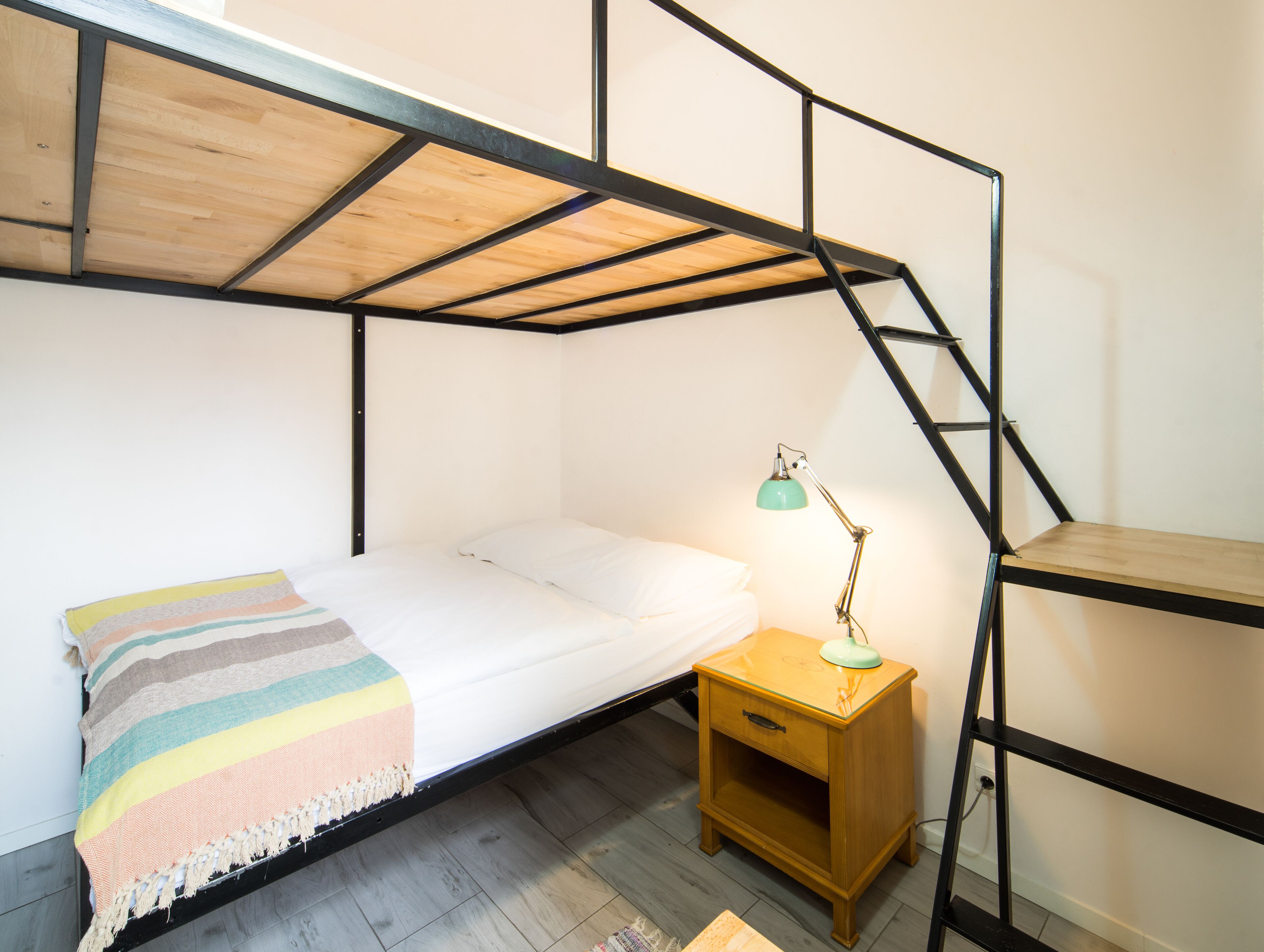Uma cama beliche com estrutura de madeira, uma mesa de cabeceira com abajur e uma pequena prateleira.