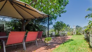 Uma área de estar coberta ao ar livre, com cadeiras vermelhas e uma mesa, rodeada por vegetação e céu azul.