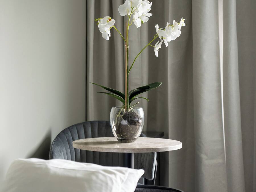 Quarto de hotel com cama, uma cadeira, uma pequena mesa redonda com um vaso de flores brancas e uma janela com cortinas.