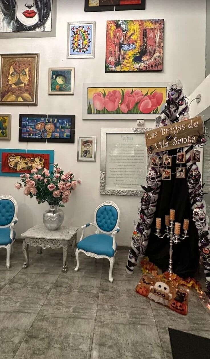 Um quarto com uma parede decorada com diversas obras de arte emolduradas, uma pequena mesa com um vaso de flores e uma exposição de caveiras e velas.