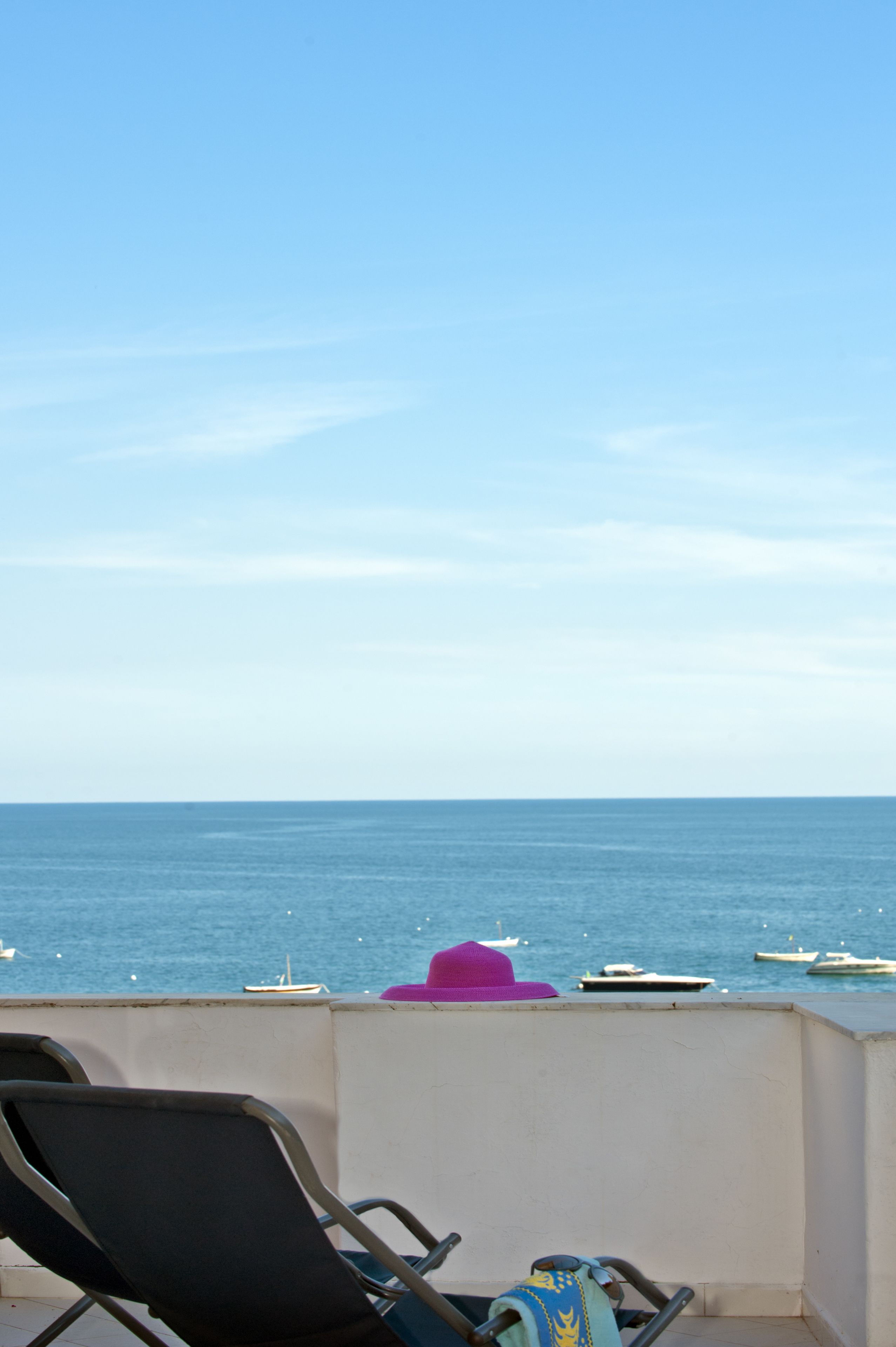 Varanda com duas cadeiras de lounge, um chapéu rosa e uma bolsa azul. Barcos podem ser vistos na água.