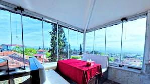 Quarto com uma cama, um banco e janelas amplas com vista para a cidade.