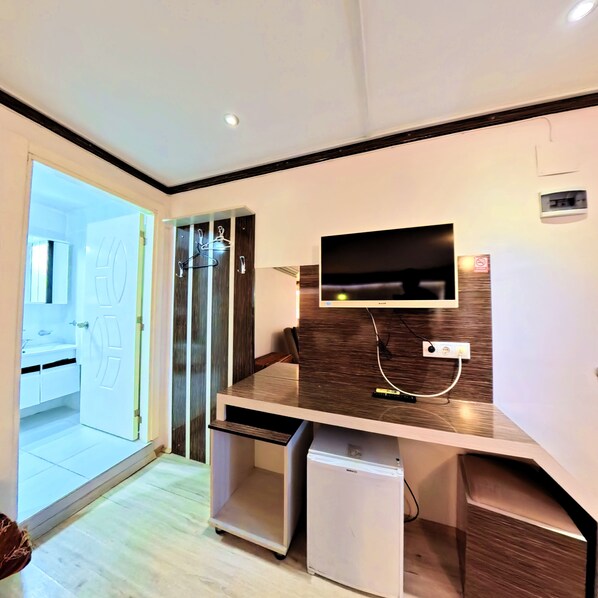 Quarto moderno com televisão embutida, mini-frigorífico, cabideiro e porta de acesso a uma casa de banho.