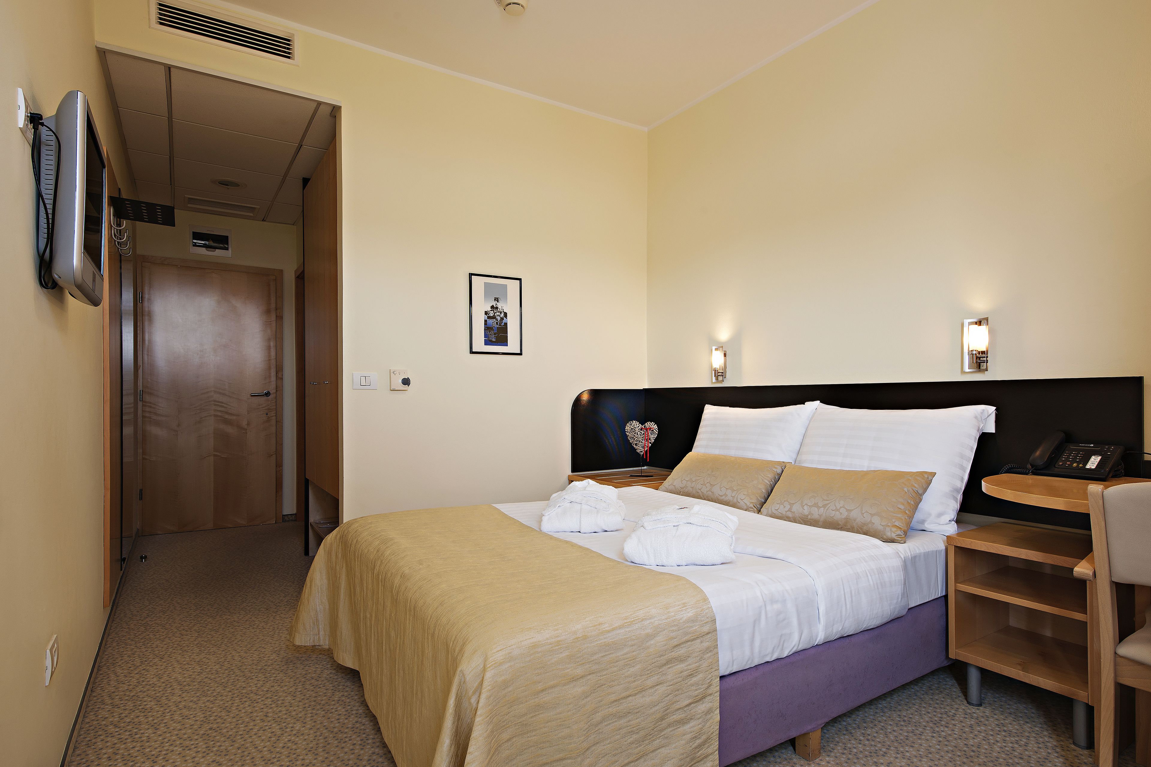 Quarto de hotel com uma cama, uma escrivaninha e uma cadeira. Há uma porta que dá para outro cômodo e um quadro na parede.