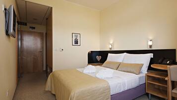 Quarto de hotel com uma cama, uma escrivaninha e uma cadeira. Há uma porta que dá para outro cômodo e um quadro na parede.
