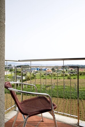 Varanda com cadeira de vime, grade de metal e vista para um campo verde e edifícios ao longe.