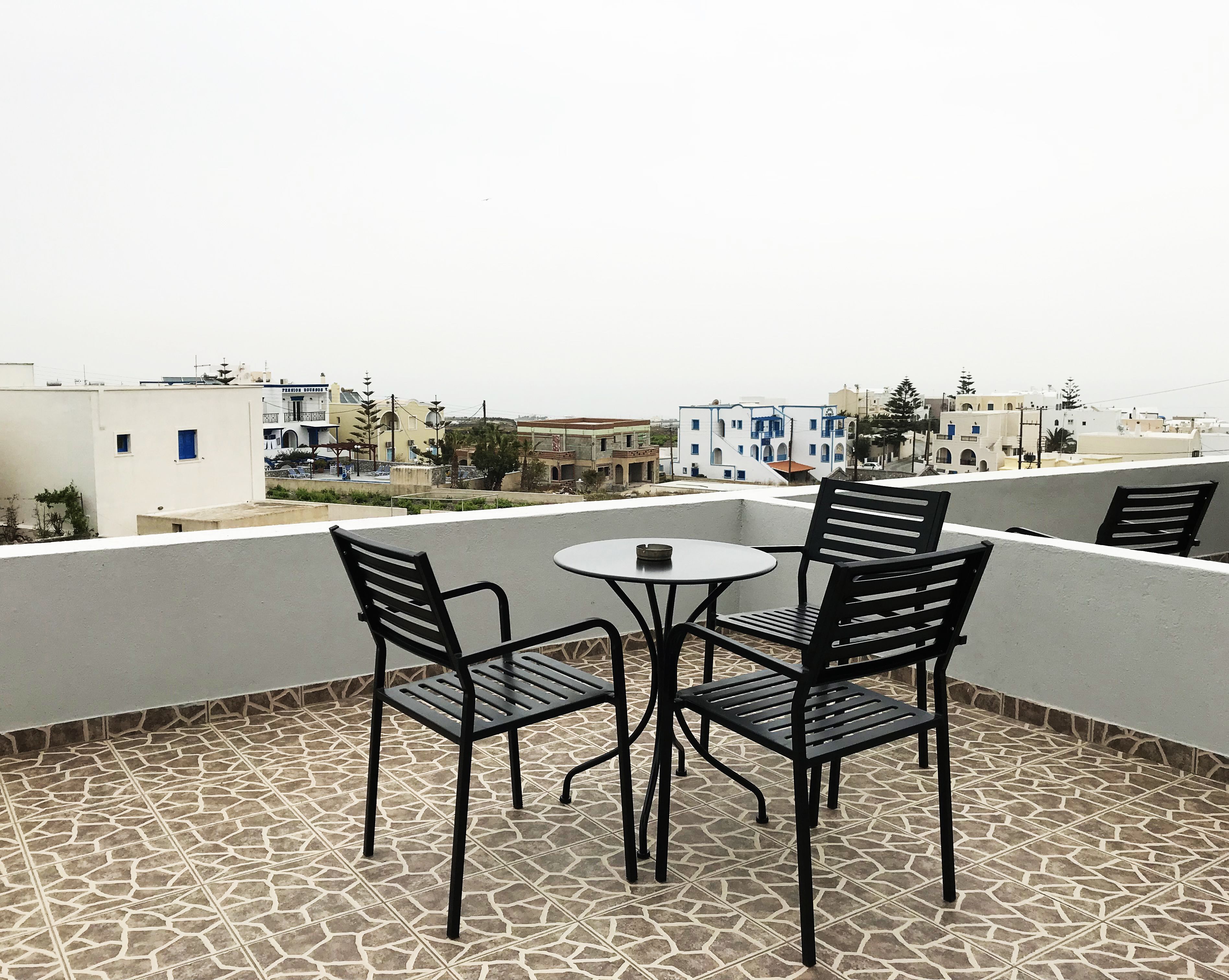 Terraço com mesa, duas cadeiras, piso estampado e edifícios ao fundo.