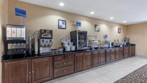 Espaço de café da manhã de hotel com estação de café à vontade, bar de sucos e refrigerador com produtos.