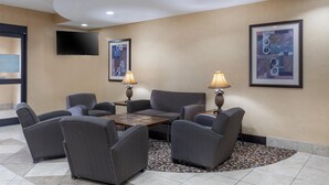 Um saguão de hotel com área de estar, incluindo um sofá, poltronas, uma mesa de centro e duas luminárias fixadas na parede. Há uma TV de tela plana na parede e obras de arte emolduradas acima da área de estar.