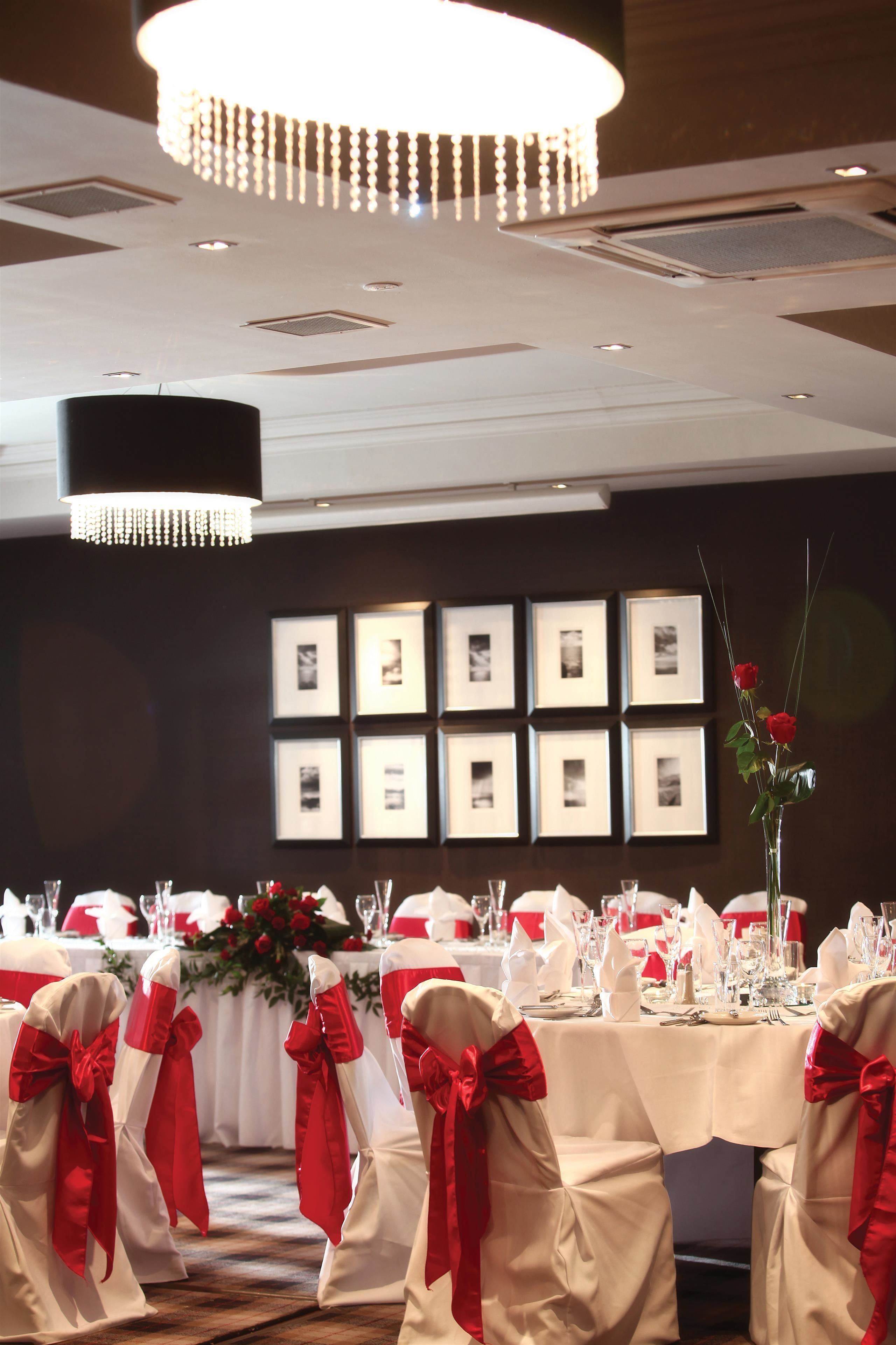 Uma sala de banquetes com mesas montadas para um evento formal, com toalhas brancas, fitas vermelhas e um lustre.
