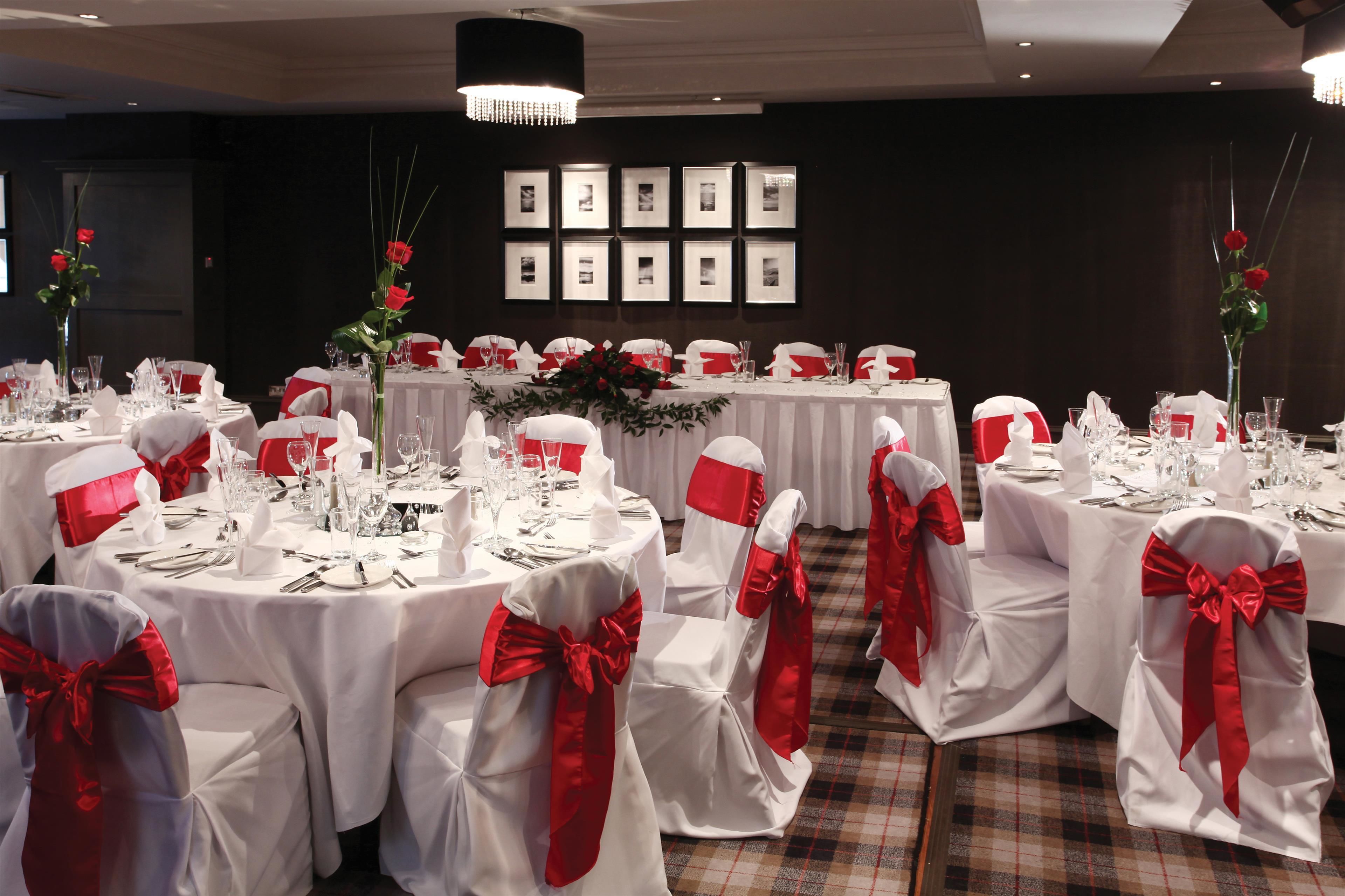 Uma sala de banquetes com mesas redondas preparadas para refeições, toalhas brancas e fitas vermelhas nas cadeiras.