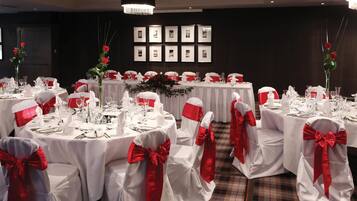 Uma sala de banquetes com mesas redondas preparadas para refeições, toalhas brancas e fitas vermelhas nas cadeiras.