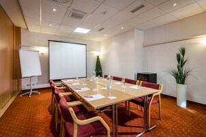 Uma sala de conferências com uma mesa comprida, cadeiras vermelhas e amarelas, um quadro branco e uma tela de projeção.