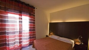 Quarto com cama, mesa de cabeceira, luminária e janela com cortinas.