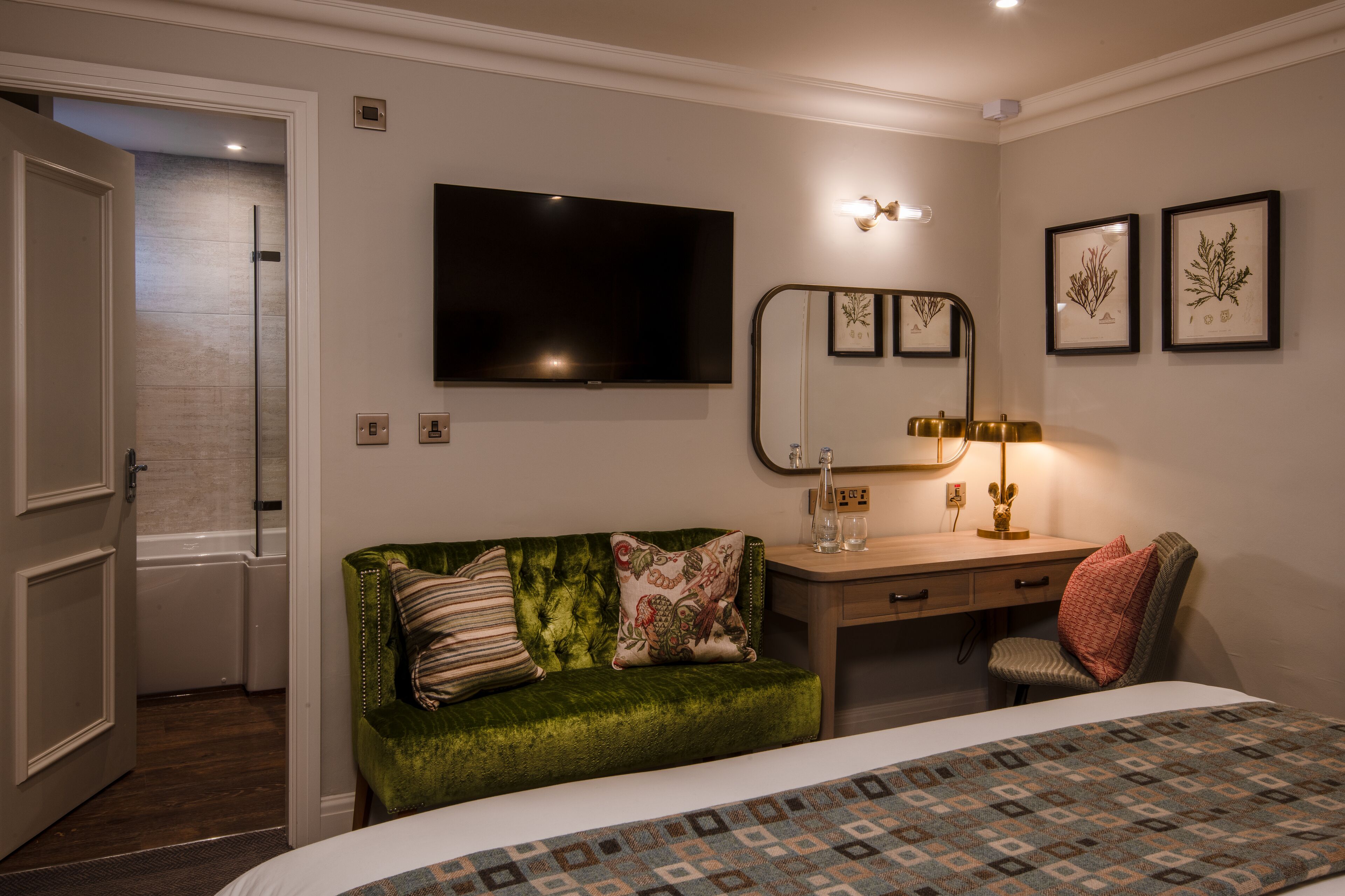 Quarto de hotel com sofá de veludo verde, escrivaninha de madeira, cama com colcha estampada, televisão de tela plana, espelho fixado na parede e gravuras botânicas emolduradas.