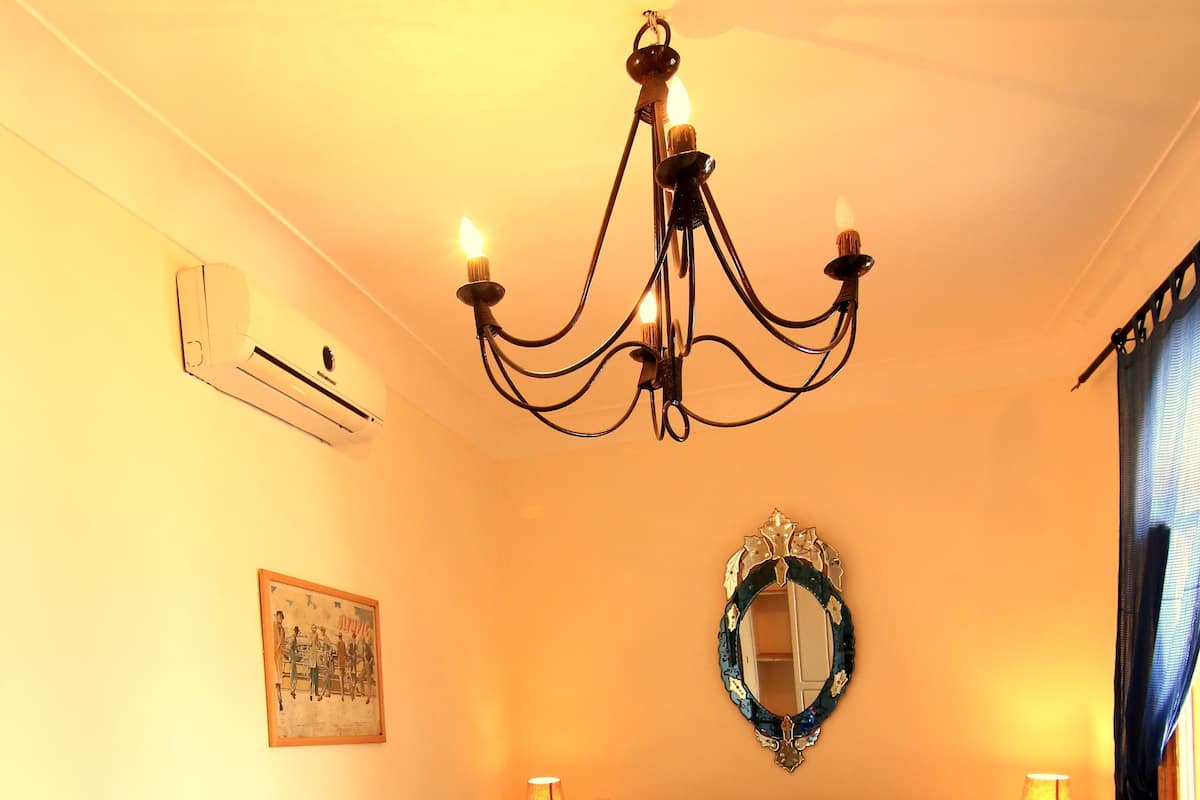 Quarto de hotel com uma cama, duas mesas de cabeceira com abajures, um candelabro, um espelho, um ar condicionado fixado na parede e uma pequena mesa com uma garrafa de água e um controle remoto.